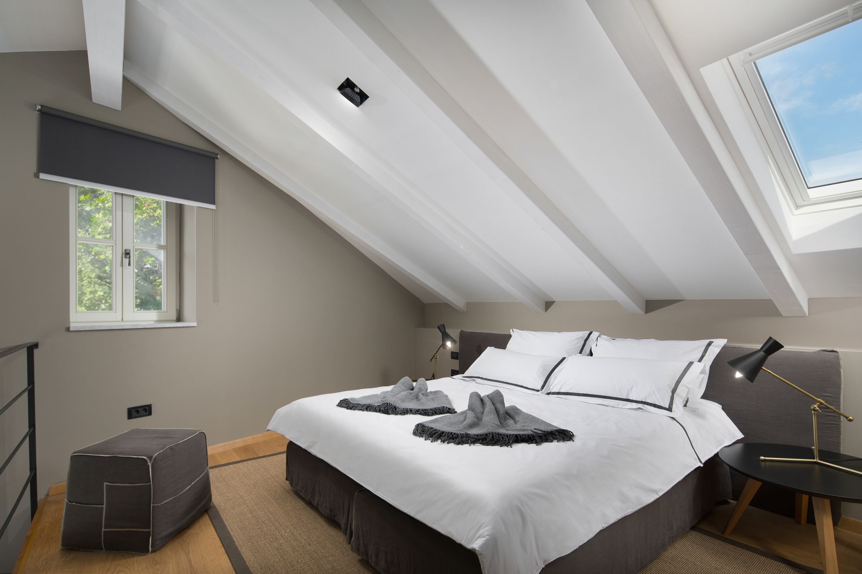 Quarto moderno com teto inclinado, uma cama grande com roupa de cama branca, uma mesa de cabeceira com abajur e uma janela com vista para o céu.