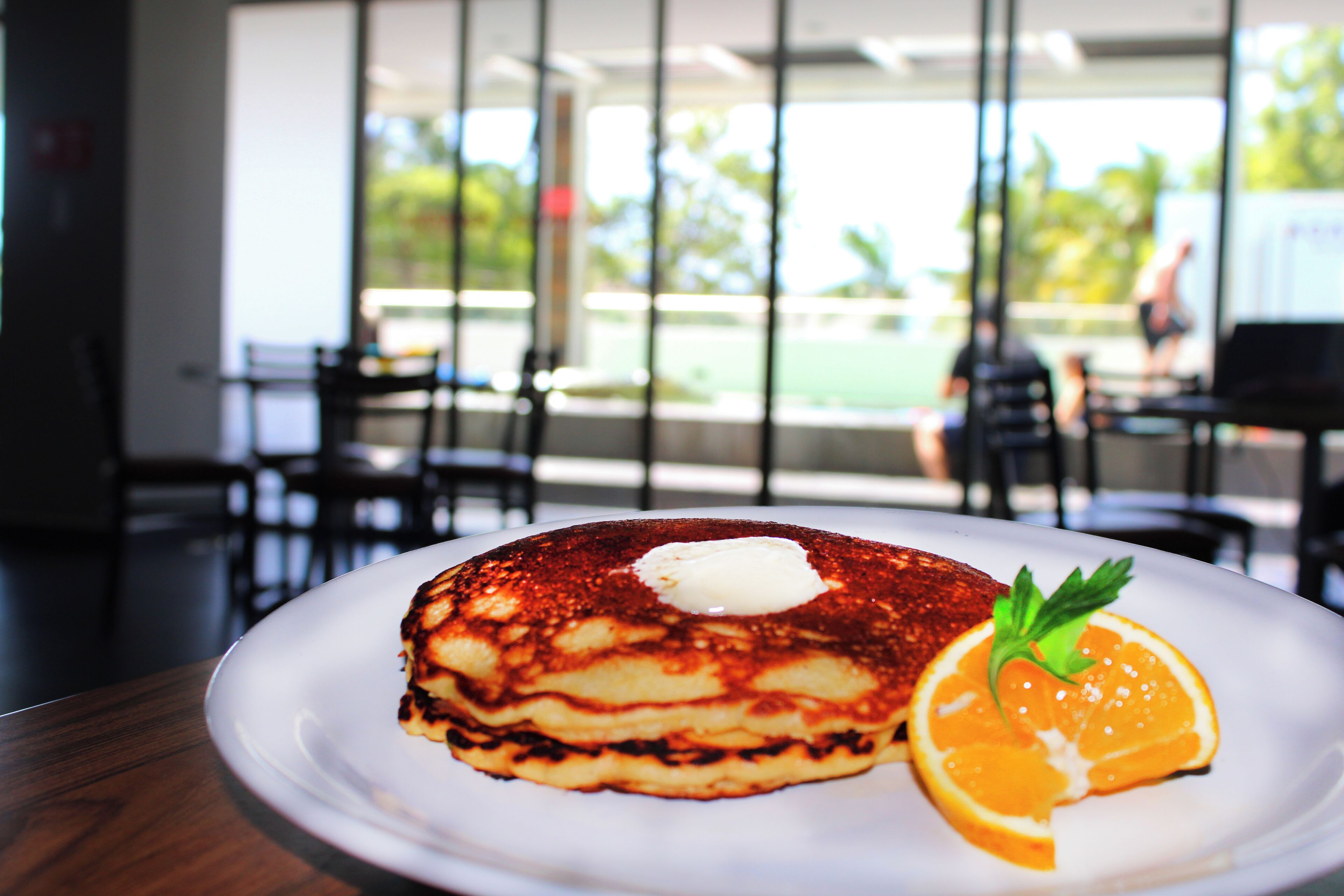 Uma pilha de panquecas com calda e uma porção de manteiga, decorada com uma fatia de laranja e um raminho de salsa, sobre um prato branco.