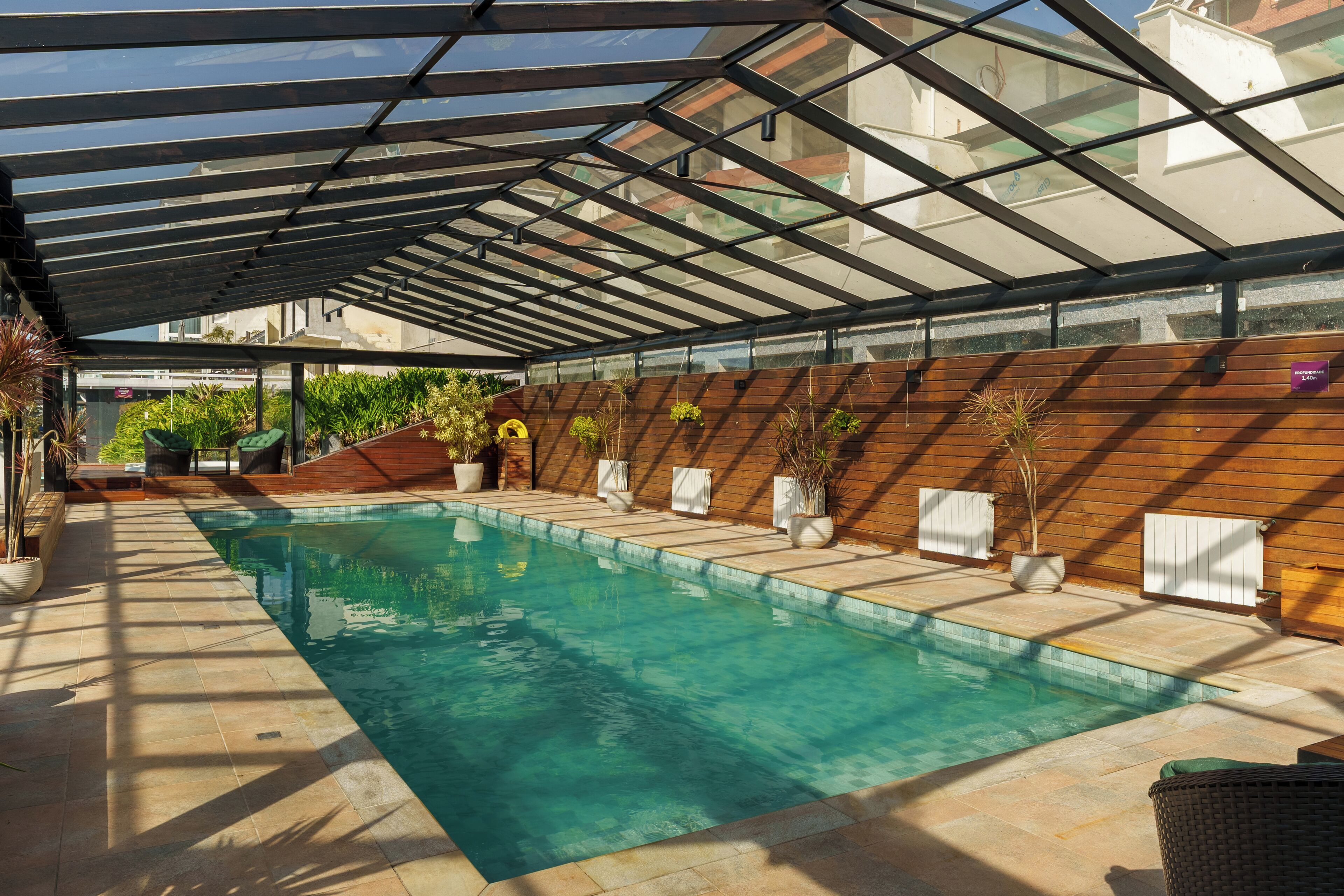 Piscina com teto de vidro, rodeada por plantas em vasos e paredes de madeira.