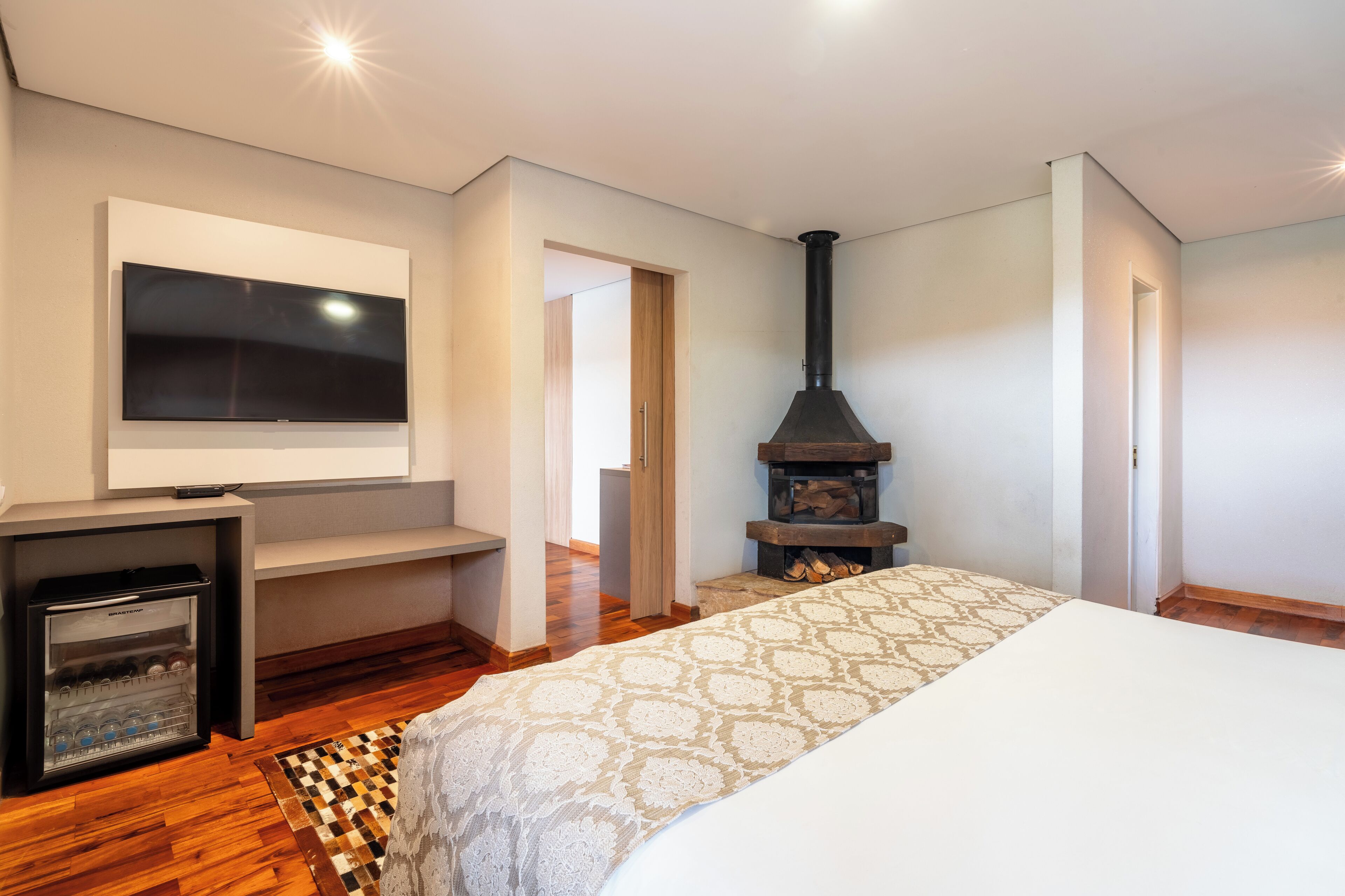 Quarto moderno com cama grande, TV integrada, lareira e pequeno frigorífico.