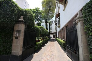 Uma estreita passagem de pedra intercalada por sebes e portões de metal preto, conduzindo a um edifício com fachada branca e um pilar de pedra com o número 31.