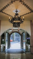 Entrada de hotel com um lustre decorativo, portas em arco e vista da rua.