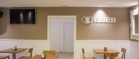 Interior de restaurante com um letreiro na parede que diz "USABARENA", mesas e cadeiras de madeira, uma TV de tela plana na parede e uma porta ao fundo.