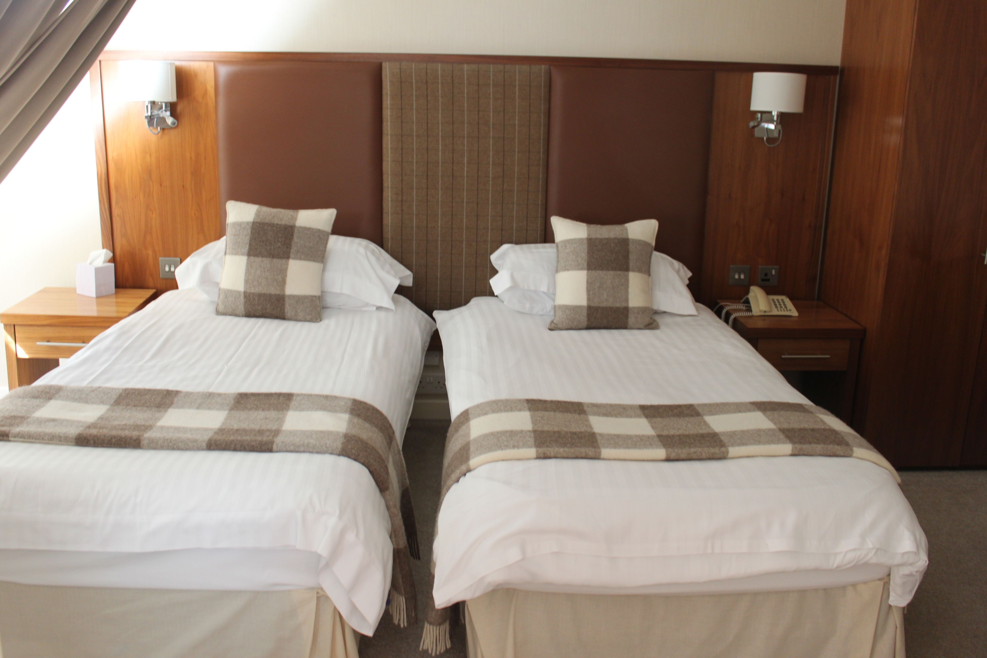 Quarto de hotel com duas camas, cada uma com manta xadrez, roupa de cama branca e cabeceira de madeira. Há abajures e telefone em cada mesa de cabeceira.