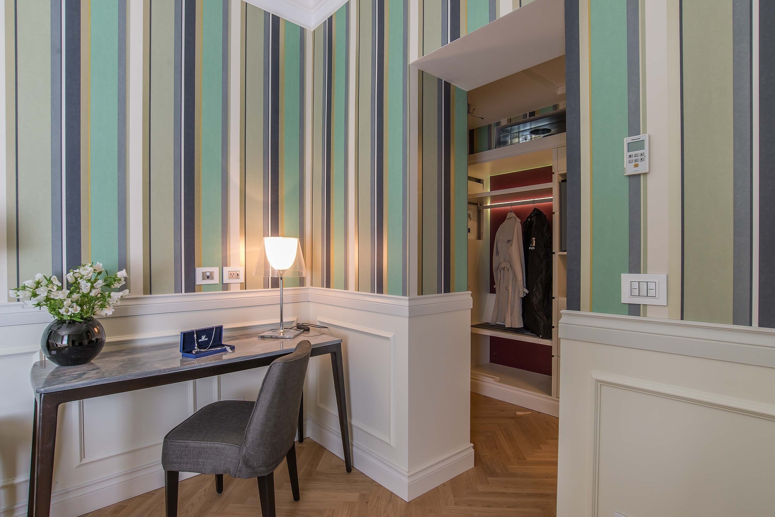 Quarto de hotel com parede listrada, uma escrivaninha com cadeira, um vaso de flores e um cabideiro com roupas.