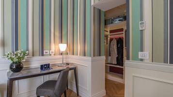 Quarto de hotel com parede listrada, uma escrivaninha com cadeira, um vaso de flores e um cabideiro com roupas.
