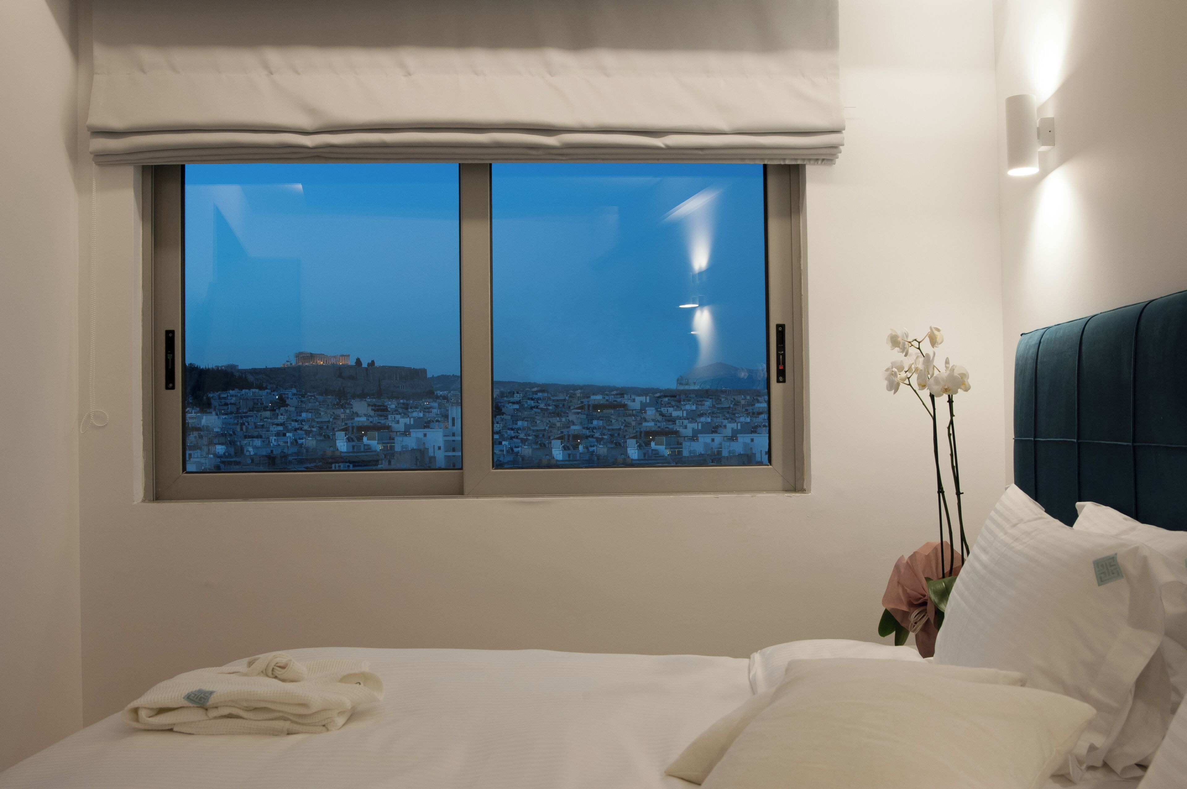 Quarto de hotel com cama, janela com vista para a paisagem urbana e luminária fixada na parede.