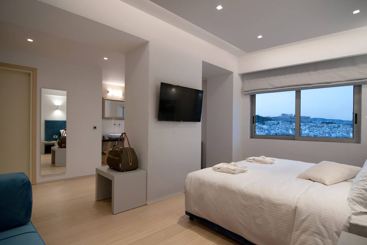 Quarto de hotel moderno com uma cama grande, televisão fixada na parede, janela com vista para a cidade e um espelho refletindo o quarto.