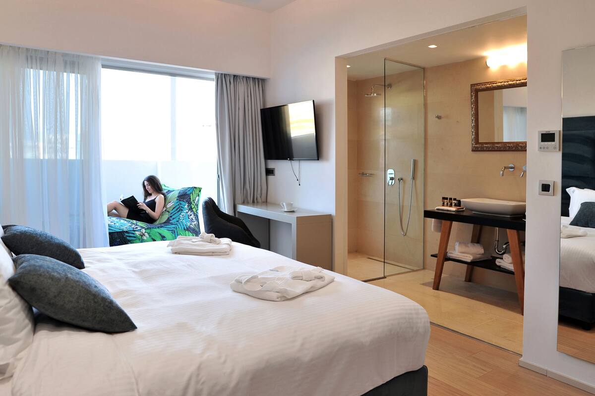 Quarto de hotel com cama grande, televisão, banheiro com chuveiro e mesa de escritório com computador.