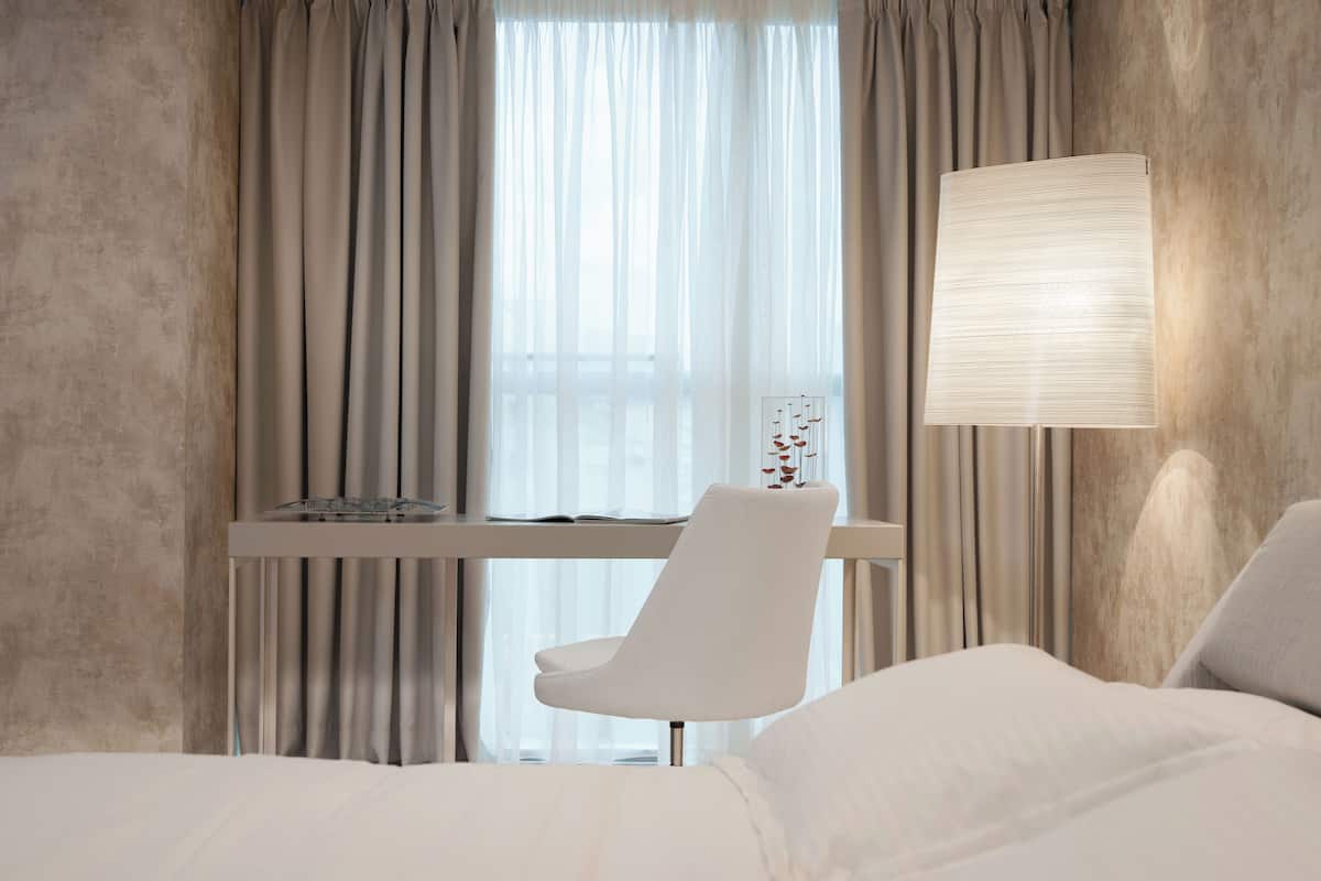 Quarto de hotel moderno com uma cama, uma cadeira branca, uma escrivaninha com abajur e cortinas.