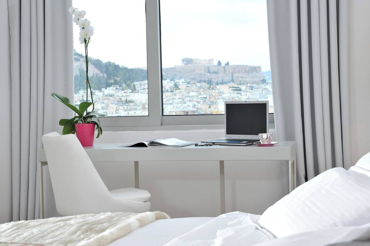 Quarto de hotel com uma cama, uma escrivaninha com um laptop, uma cadeira, uma orquídea em vaso e vista para a cidade através de grandes janelas.