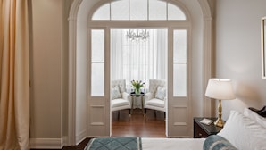 Quarto de hotel com uma grande porta arqueada que dá para uma sala de estar com duas cadeiras e uma mesa, um candelabro e um abajur.