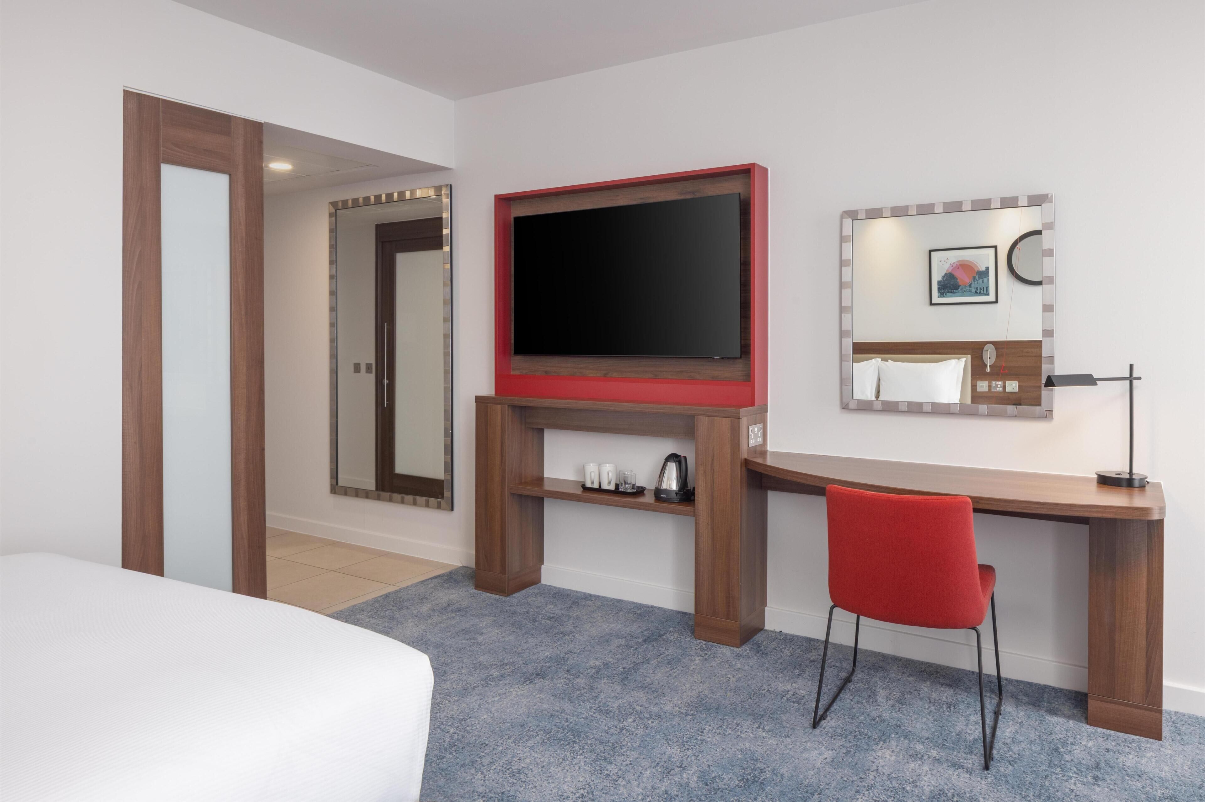 Quarto de hotel com uma cama, uma escrivaninha com cadeira, televisão, espelho e prateleira com chaleira.