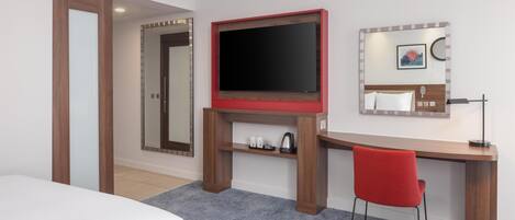 Quarto de hotel com uma cama, uma escrivaninha com cadeira, televisão, espelho e prateleira com chaleira.