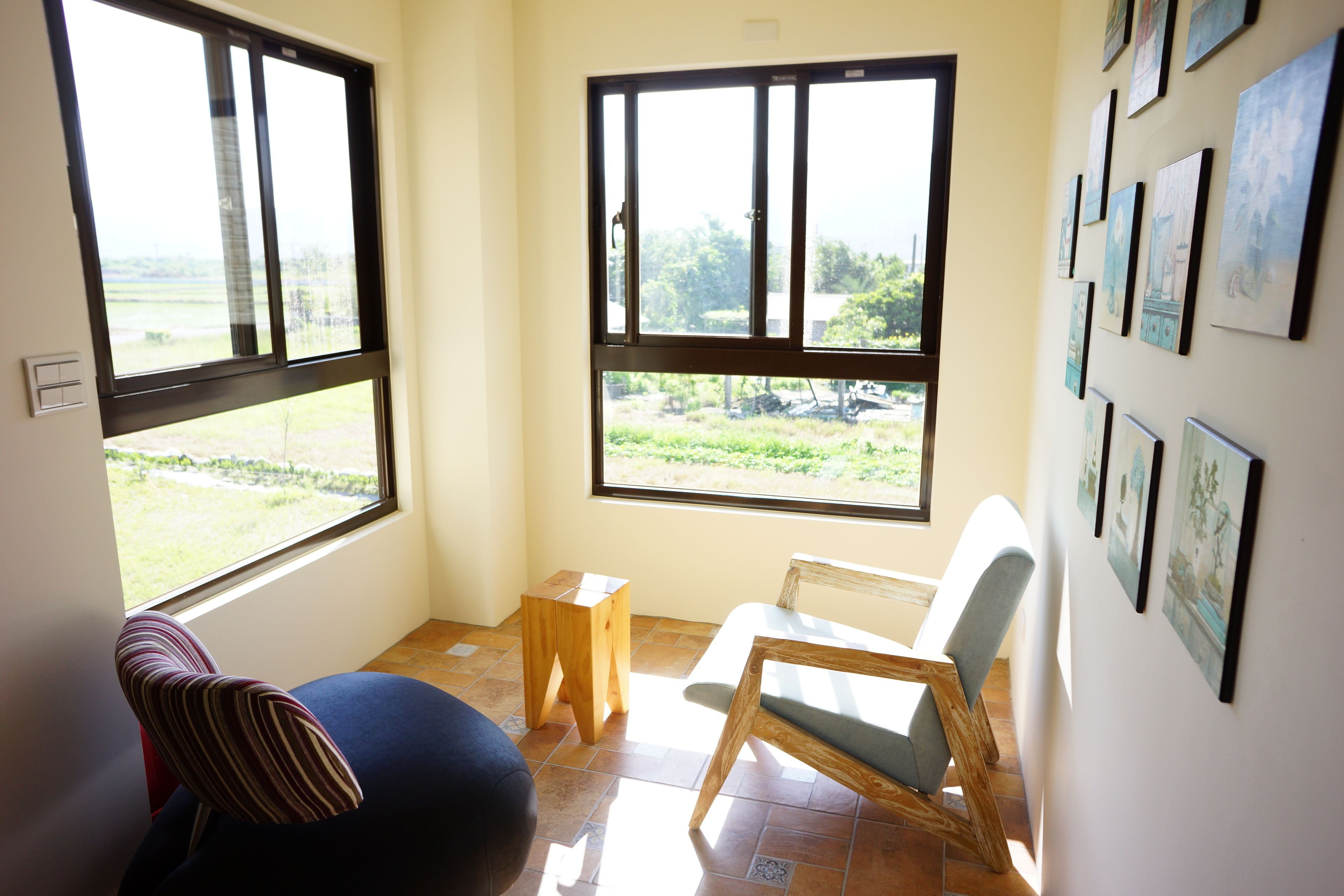 Quarto com duas janelas amplas, um banquinho de madeira, uma cadeira azul e uma cadeira branca.