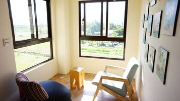 Quarto com duas janelas amplas, um banquinho de madeira, uma cadeira azul e uma cadeira branca.