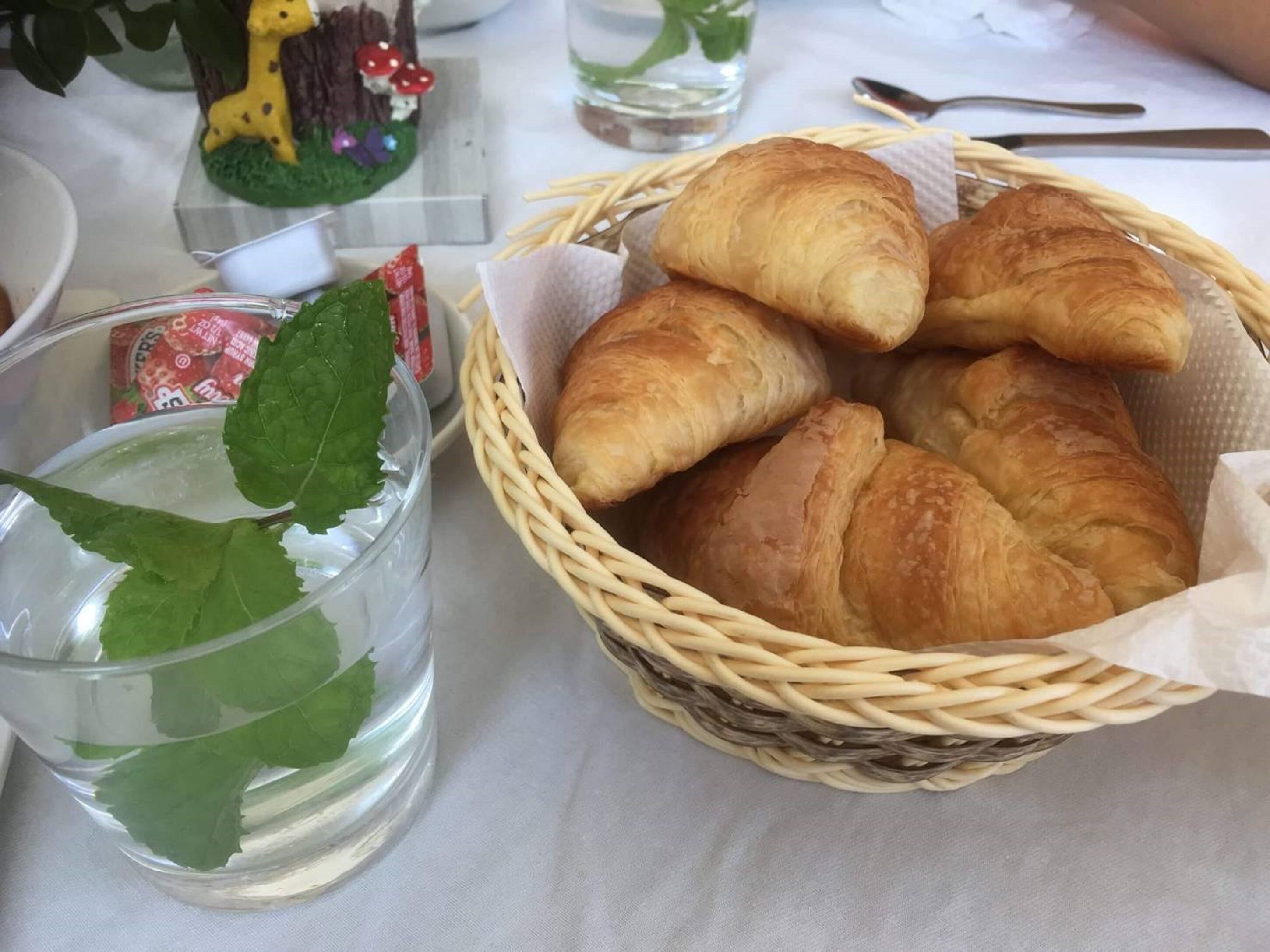 Uma cesta de croissants sobre uma mesa, com um copo de água e folhas de hortelã.