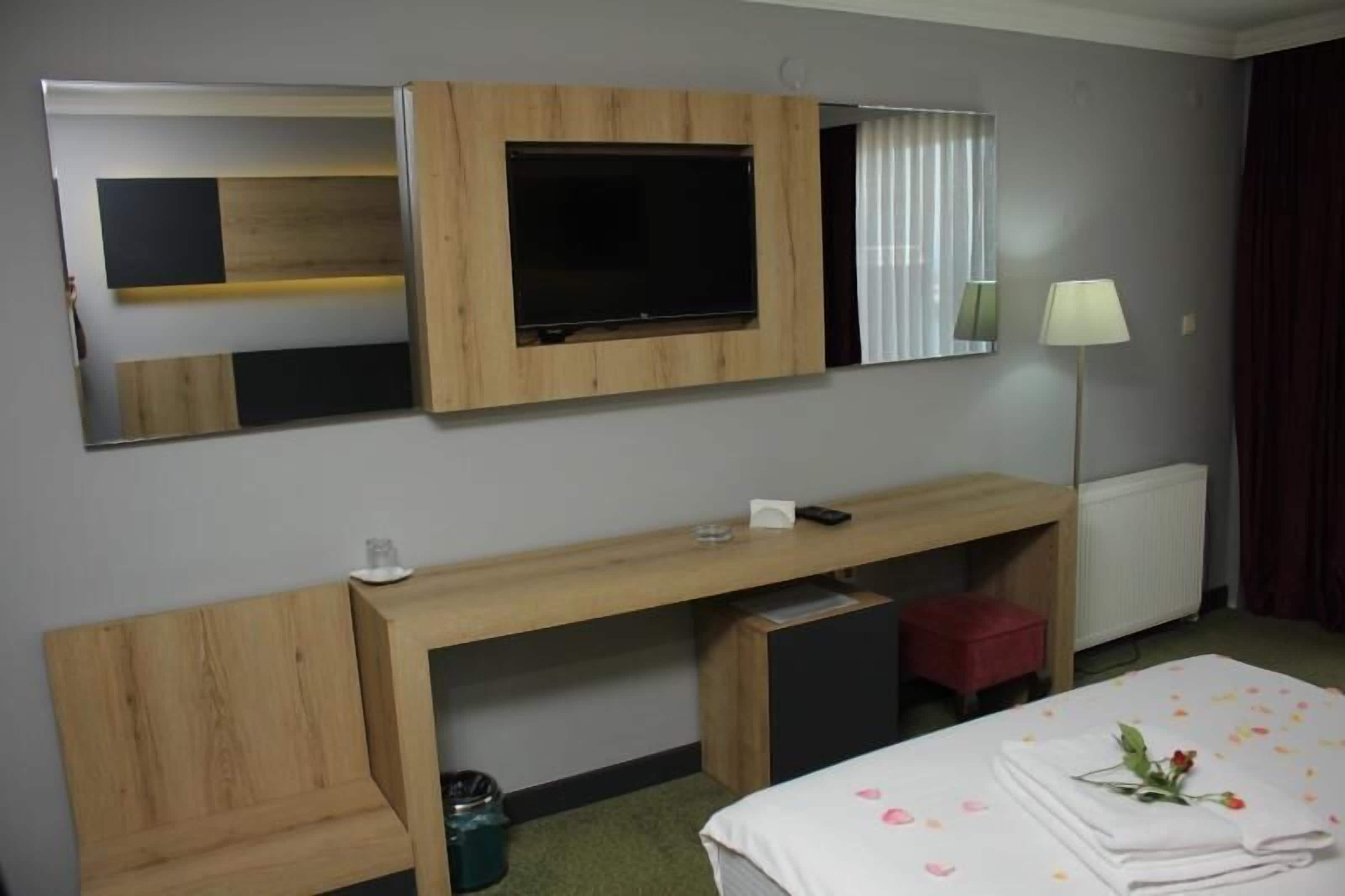 Quarto de hotel com secretária de madeira, televisão fixada na parede, luminária, cama com colcha florida e radiador.