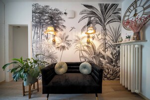 Uma sala de estar com um sofá de veludo escuro, duas luminárias fixadas na parede e um mural tropical.