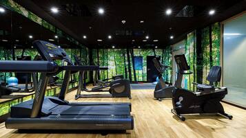 Academia com esteiras, elípticos e bicicletas ergométricas, piso de madeira e painel de parede com uma parede verde e florida.