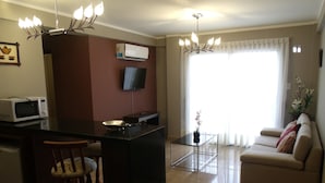 Uma sala de estar moderna com sofá, área de jantar com balcão, televisão de tela plana e uma janela grande com cortinas.