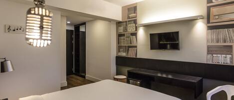 Quarto de hotel moderno com uma cama grande, uma escrivaninha, uma cadeira, uma TV fixada na parede e uma estante com diversos objetos.