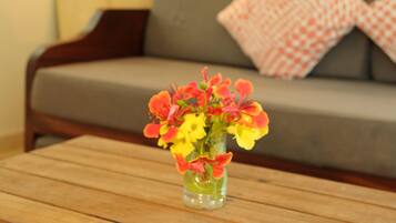 Uma mesa de madeira com um vaso de vidro de flores, uma prancha de surf e um sofá com almofadas estampadas.