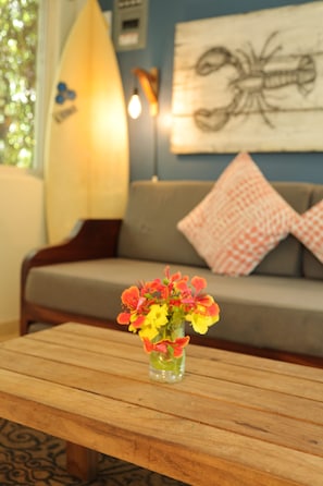 Uma mesa de madeira com um vaso de vidro de flores, uma prancha de surf e um sofá com almofadas estampadas.