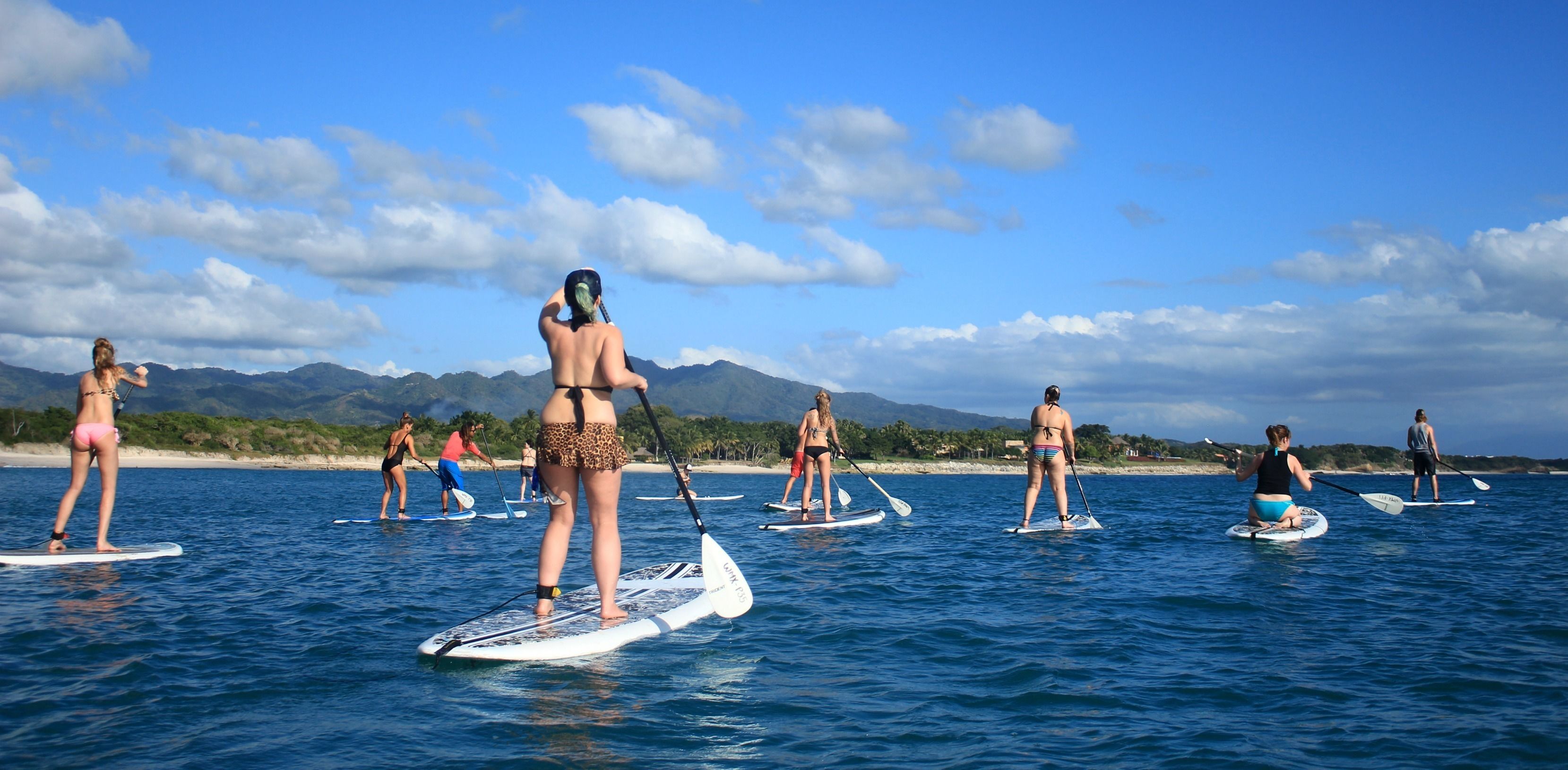 Pessoas praticando paddleboard em um mar calmo, com montanhas ao fundo.