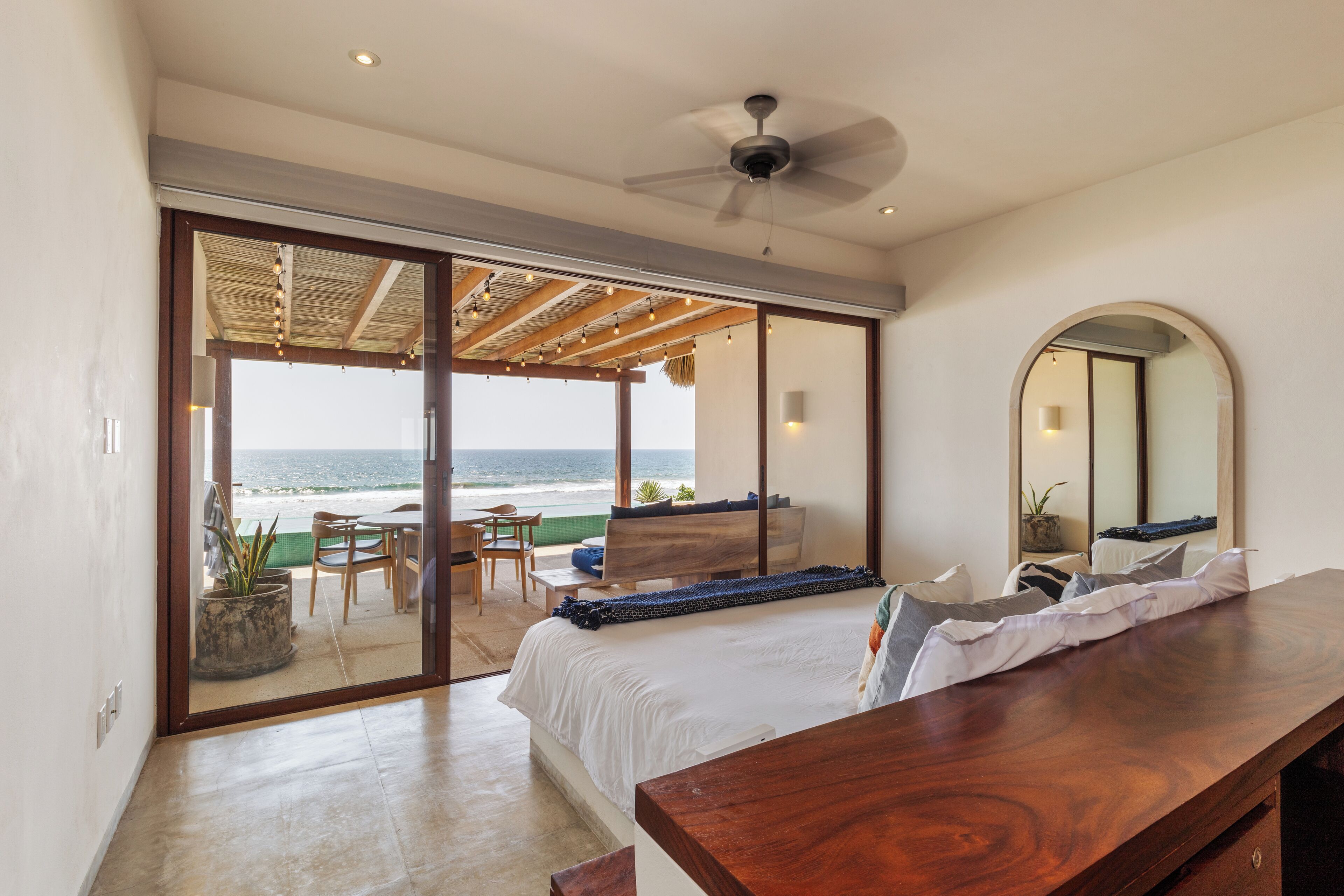 Quarto com uma cama grande, uma escrivaninha de madeira, um ventilador de teto e uma porta de vidro corrediça que dá para uma varanda com vista para o mar.