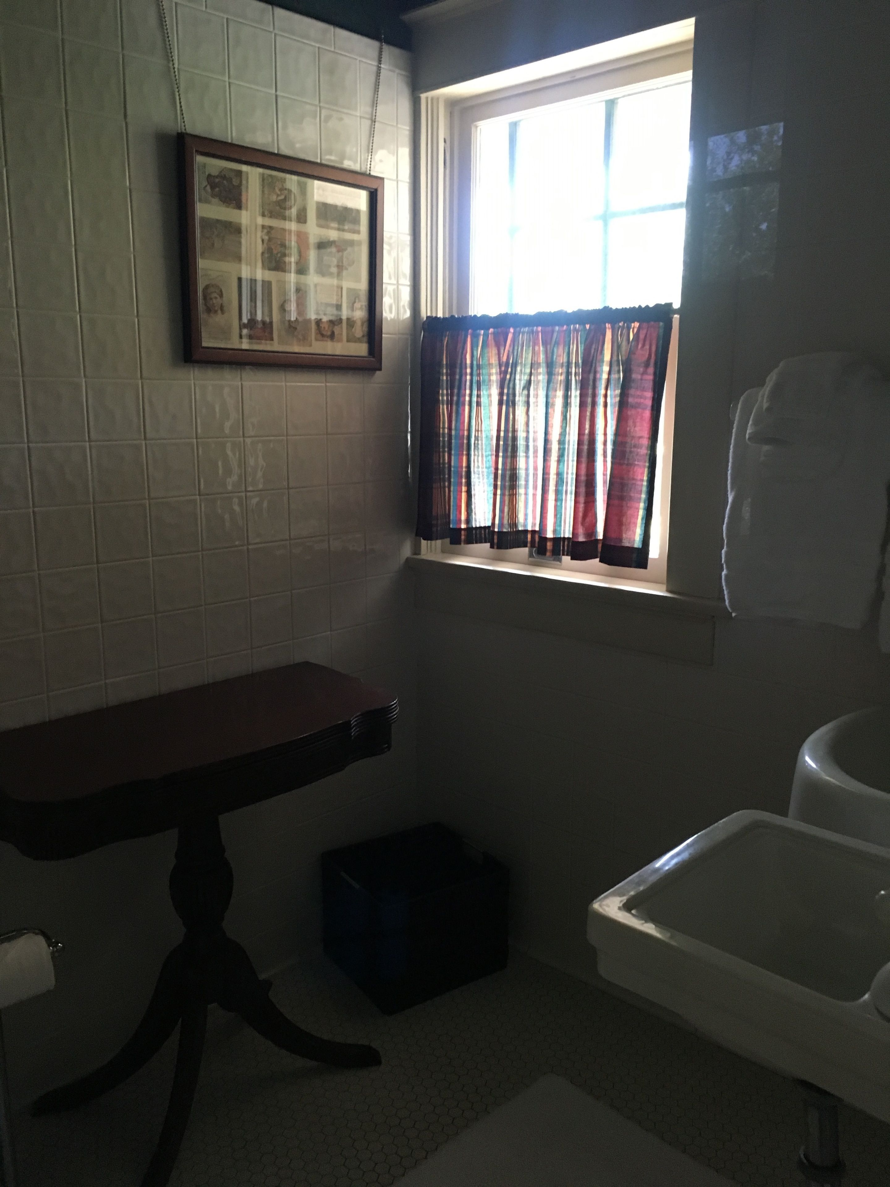 Um banheiro com parede revestida de azulejos, uma janela com cortinas coloridas, uma pia branca e um banco escuro.