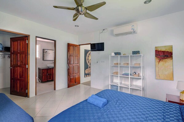 Quarto de hotel com duas camas, ventoinha no teto, televisão, estante de livros e um quadro.