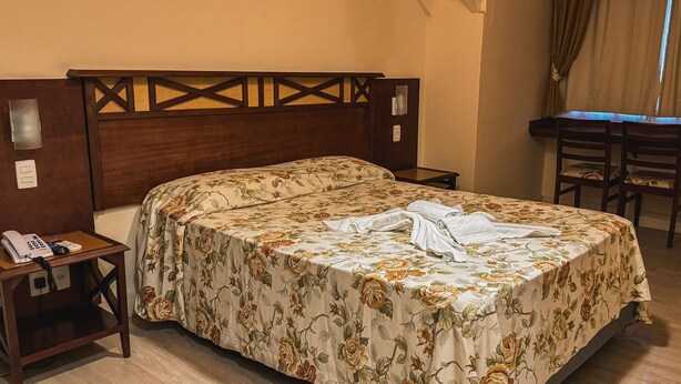 Quarto de hotel com uma cama, uma escrivaninha e uma cadeira. Há um telefone na mesinha de cabeceira.