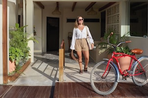 Uma pessoa em pé ao lado de uma bicicleta vermelha com cesto, em área de entrada bem iluminada com plantas em vasos e piso de azulejos.