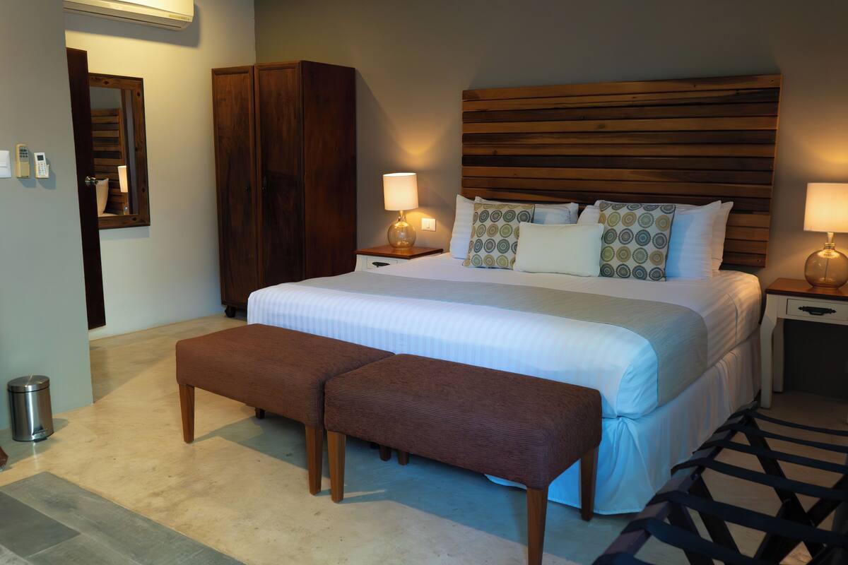 Quarto bem arrumado com uma cama grande, duas mesas de cabeceira com luminárias, um guarda-roupa de madeira e um banco.