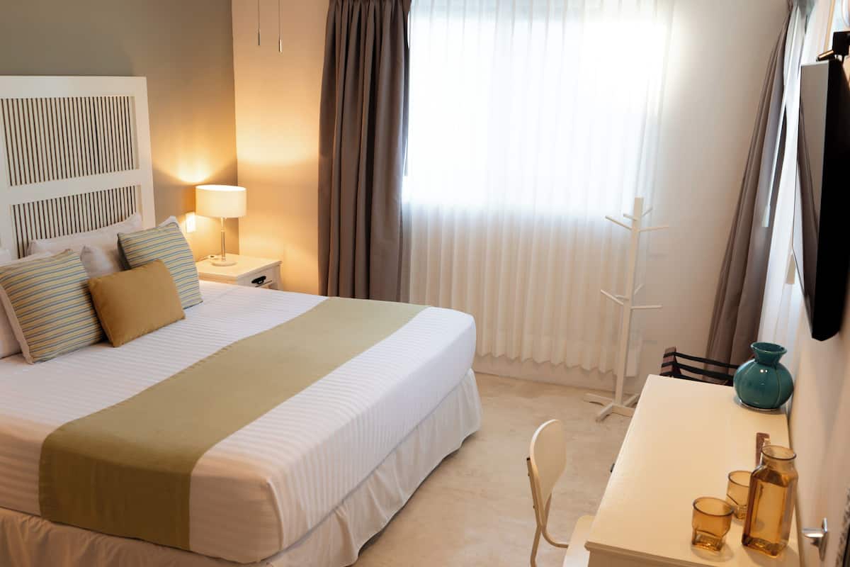 Quarto de hotel com uma cama grande, mesa de cabeceira com abajur, escrivaninha com um vaso e copos, e uma cadeira.
