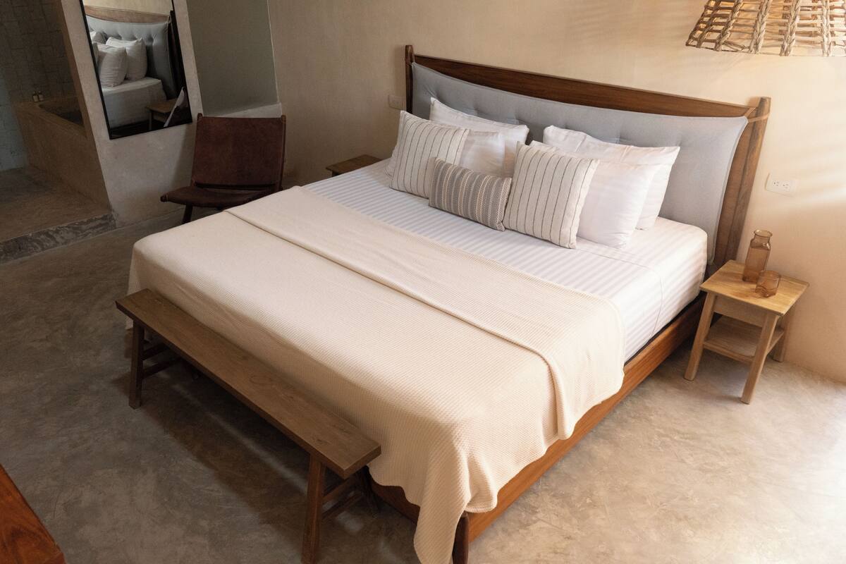 Uma cama bem arrumada com cabeceira de madeira, um banco no pé da cama e uma pequena mesa de cabeceira com uma garrafa.