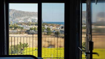 Quarto com uma grande janela com vista para a paisagem costeira, uma varanda com grade de metal e uma cama com cabeceira branca.
