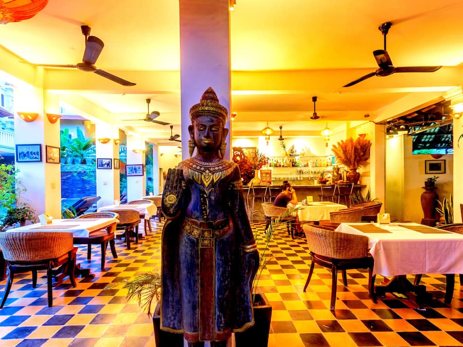 Restaurante com uma estátua de Buda como peça central, piso xadrez e plantas dentro.