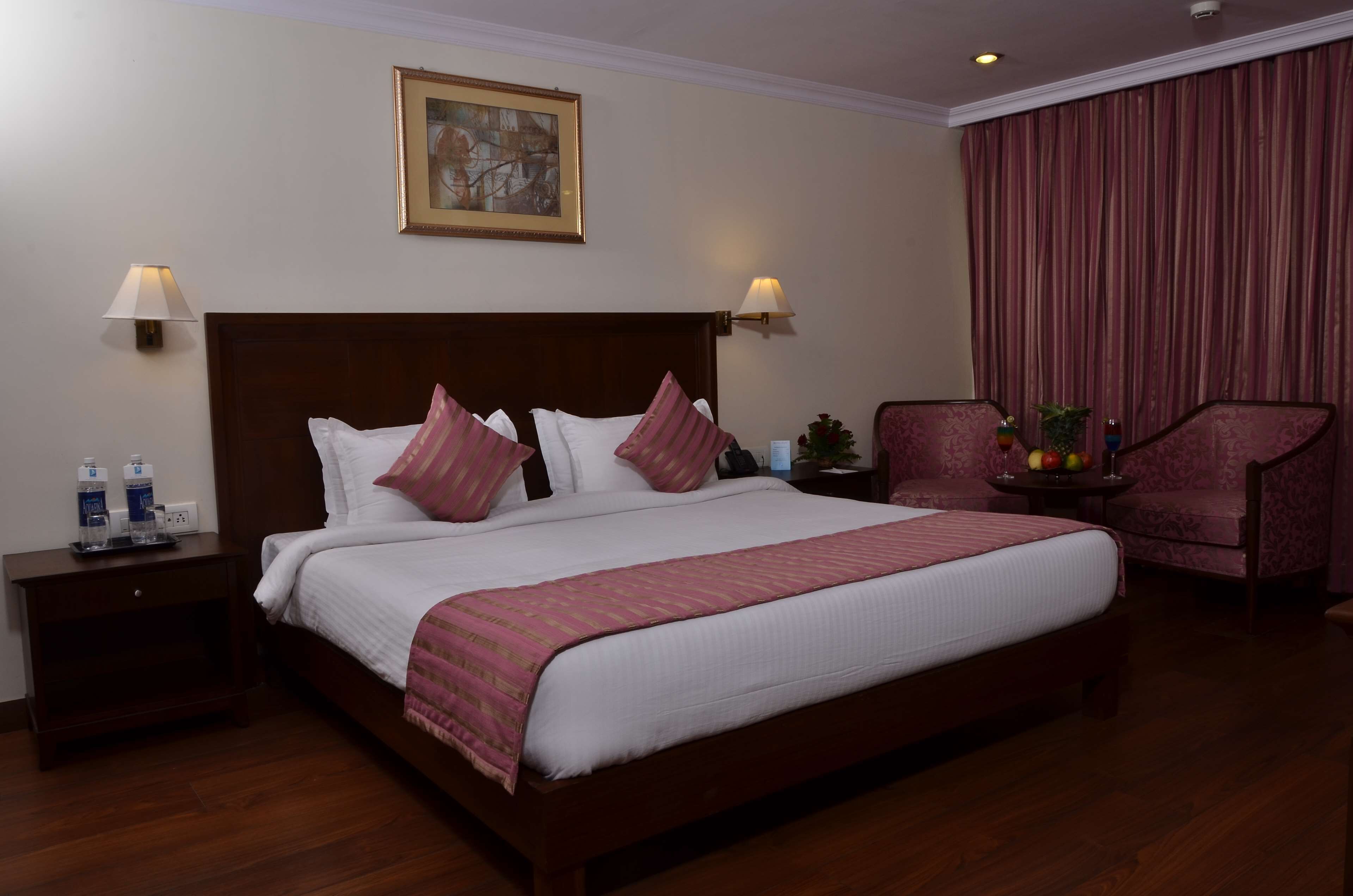Quarto de hotel com uma cama grande, duas mesas de cabeceira, uma cadeira, um quadro na parede e uma cortina.