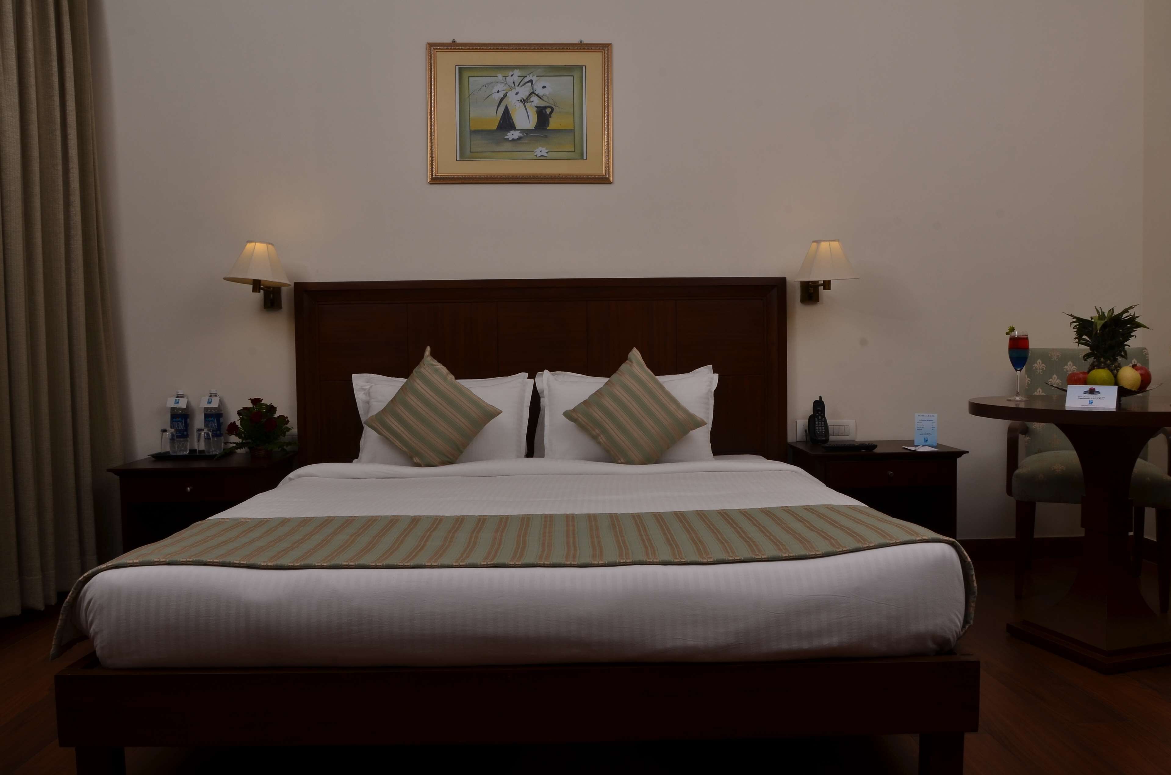 Quarto de hotel com uma cama grande, duas mesas de cabeceira, um criado-mudo, uma pequena mesa com frutas e um quadro na parede.