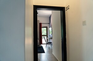 Corredor que leva a um quarto com escrivaninha e cadeira, janela com cortinas e porta com o número 202.