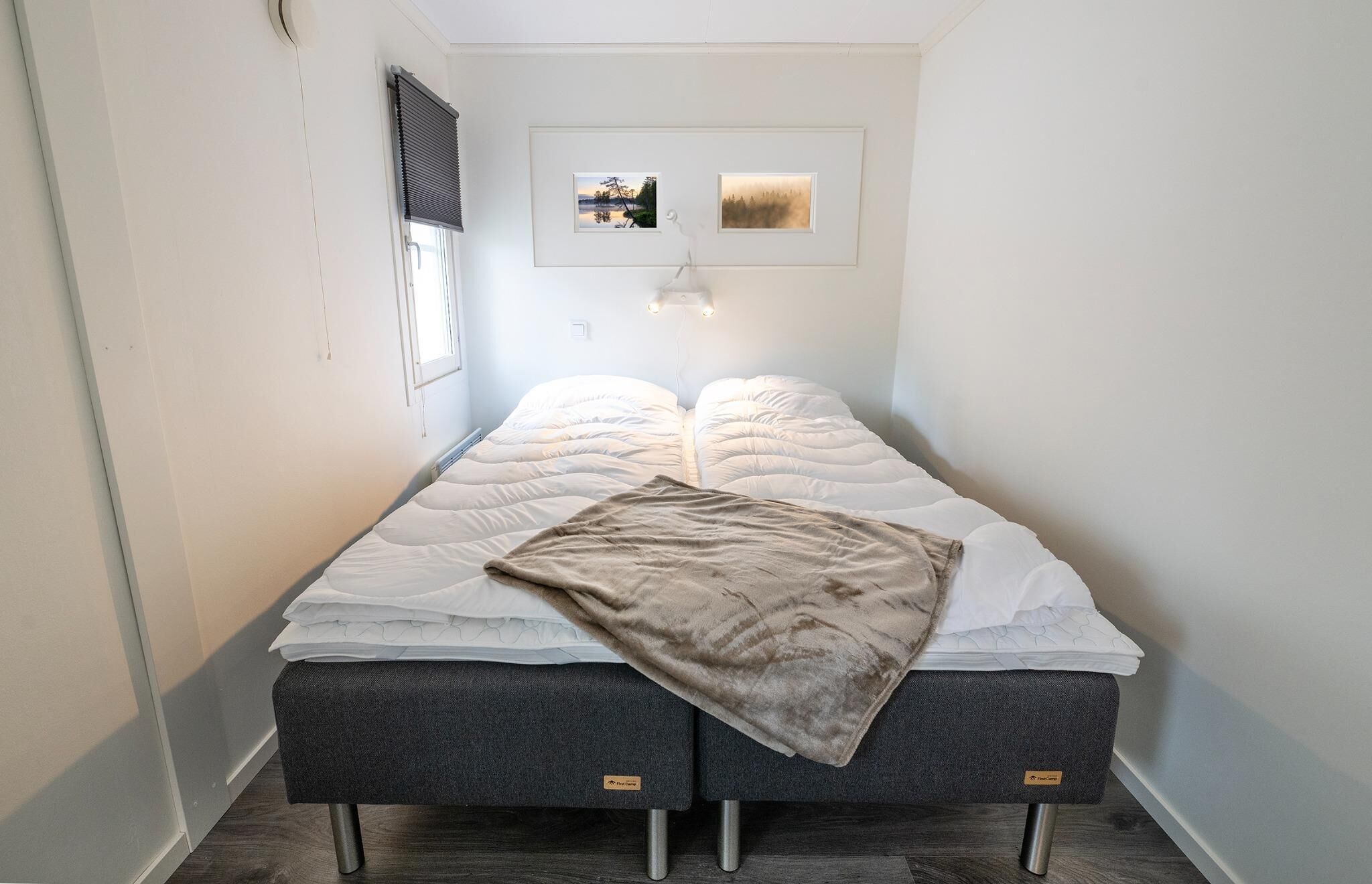 Um quarto pequeno e estreito com uma cama, uma janela com persianas e duas molduras na parede.