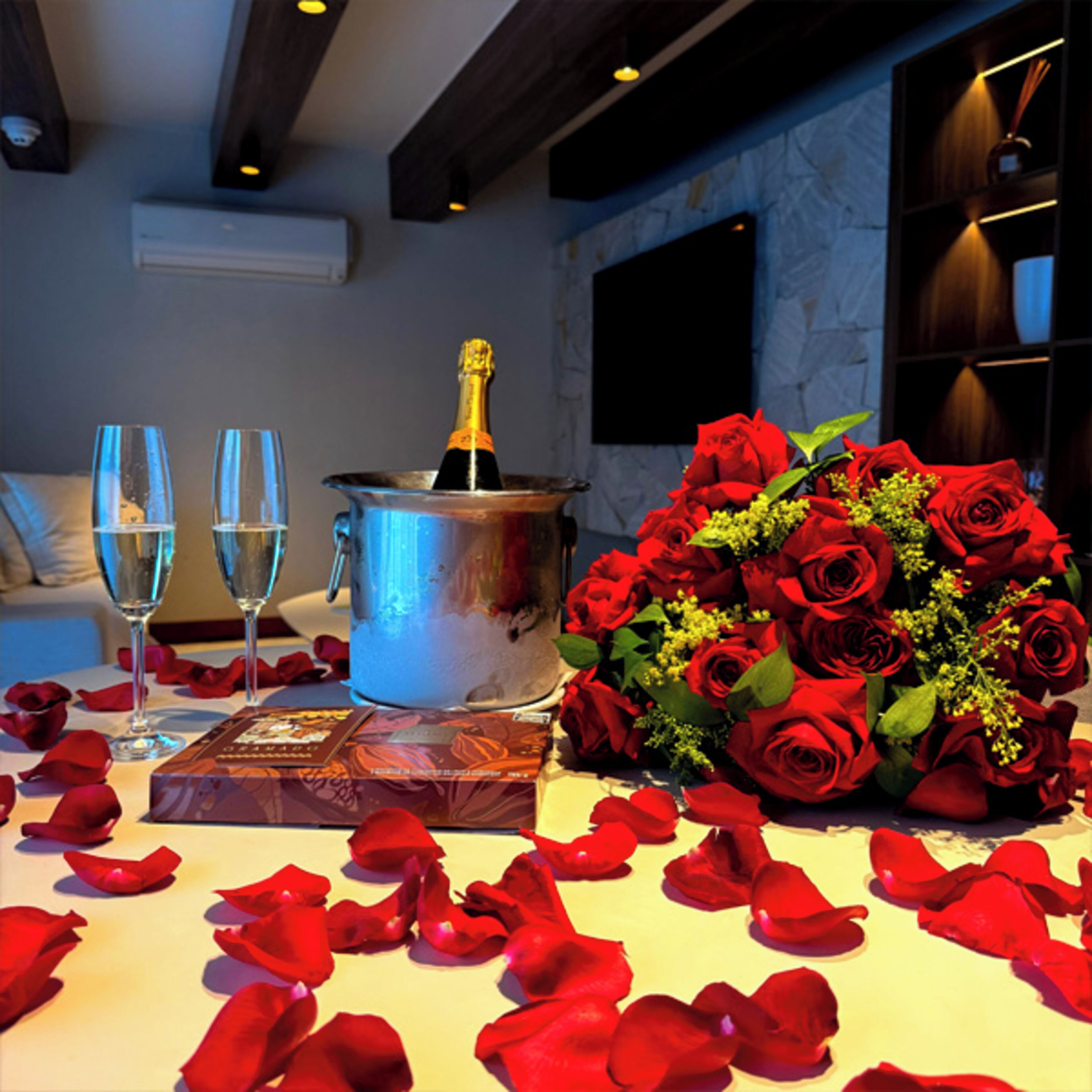 Um quarto com mesa posta para uma ocasião romântica, com champanhe, rosas e pétalas de rosa espalhadas.
