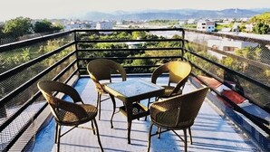 Varanda com cadeiras de vime e uma mesa, com vista para uma paisagem urbana com áreas verdes e edifícios.