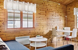 Interior de uma cabana de madeira com uma mesa de jantar, cadeiras, um sofá e uma janela com cortinas.