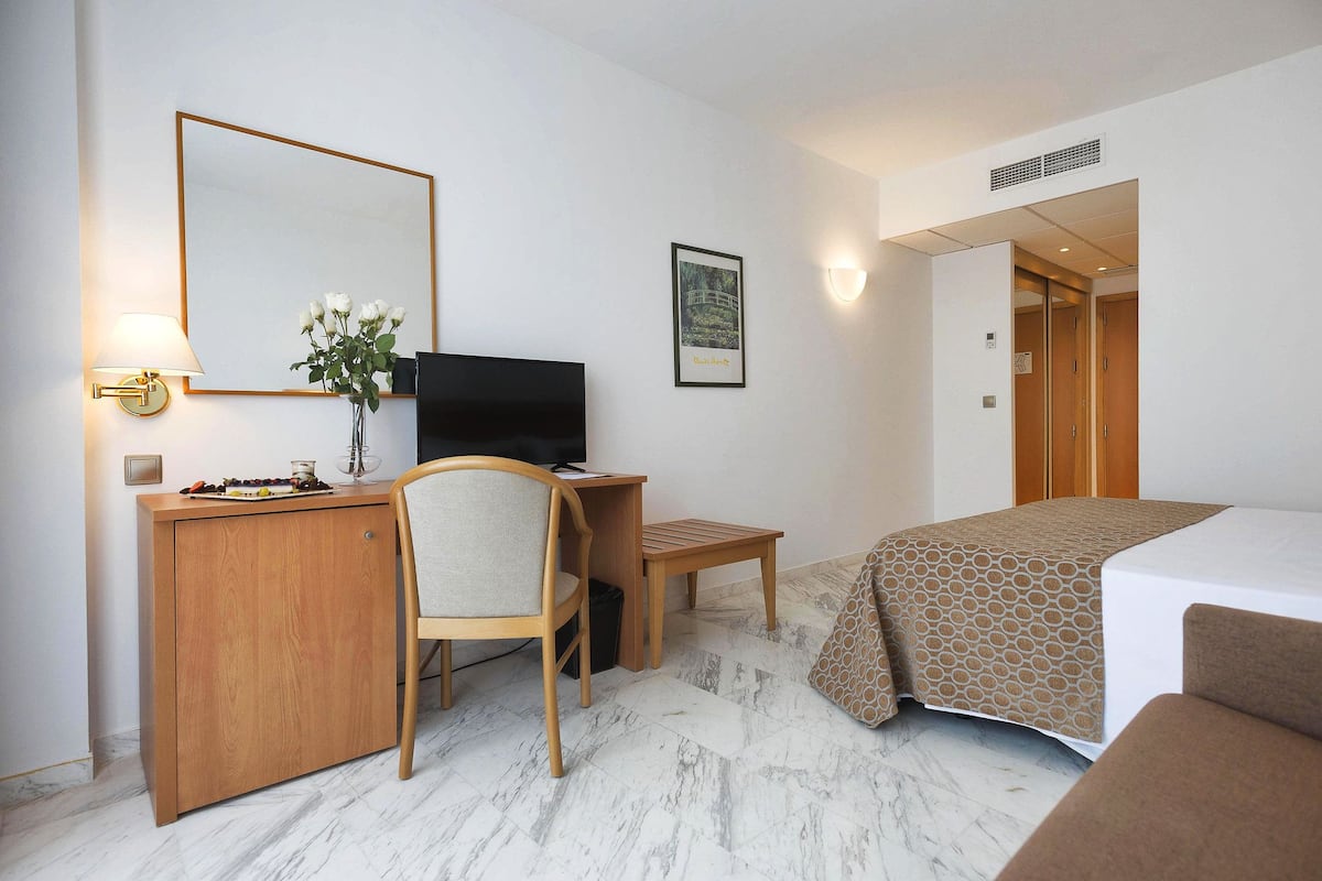 Quarto de hotel com cama, escrivaninha e cadeira, televisão, mesinha, vaso com flores, quadro na parede e guarda-roupa.