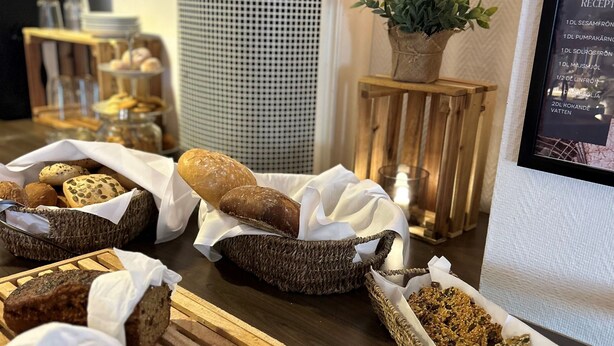Uma mesa de madeira com diversos pães e doces, uma faca e uma pequena planta em uma mesinha lateral.