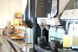Uma máquina de café a servir espresso numa chávena, com um copo de sumo de laranja e um tabuleiro de pastelaria ao fundo.