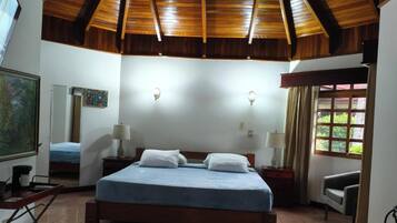 Quarto com teto de madeira, uma cama, mesas de cabeceira, uma cadeira, janela com cortinas e um quadro na parede.